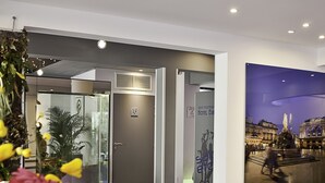 Um lobby de hotel moderno com entrada por porta de vidro, uma grande fotografia emoldurada na parede e um vaso de flores sobre uma mesa.