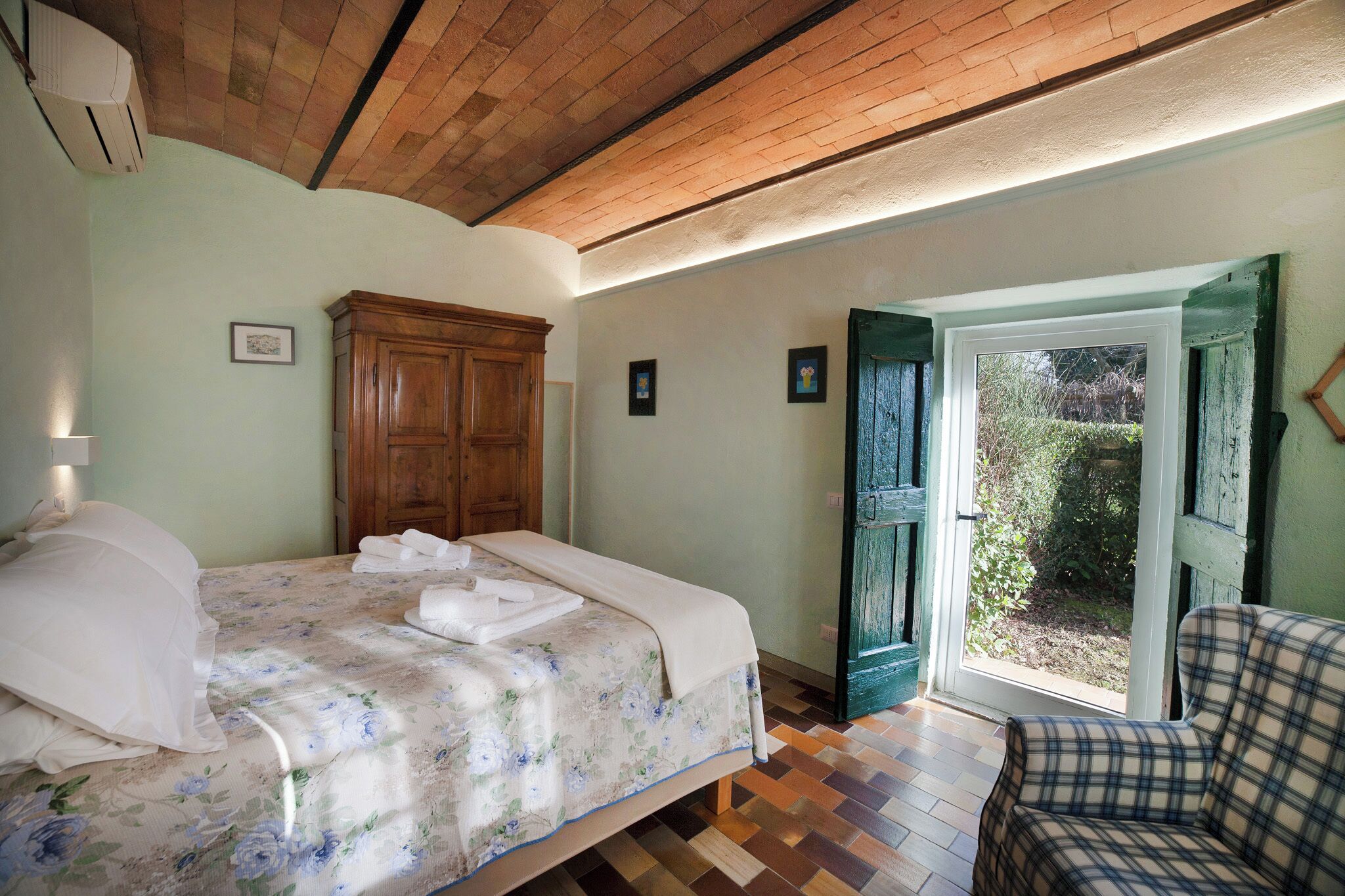 Quarto com cama, guarda-roupa de madeira, cadeira xadrez e porta para um jardim.