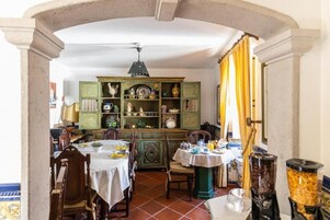 Uma sala de jantar com duas mesas, cadeiras, um armário verde com louças e um ventilador de teto.