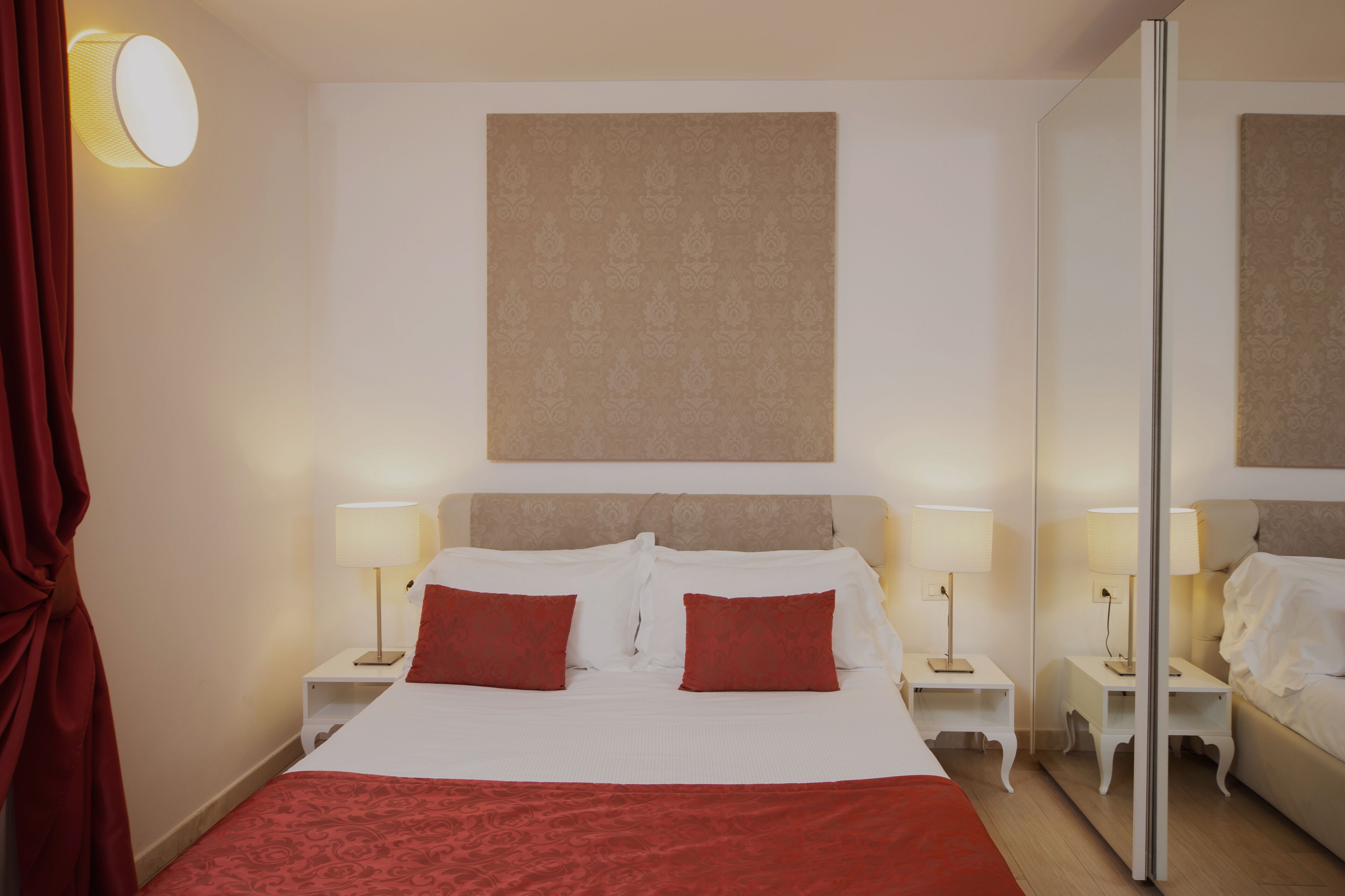 Quarto de hotel com uma cama grande, duas mesas de cabeceira com abajures, um espelho e uma cortina vermelha.