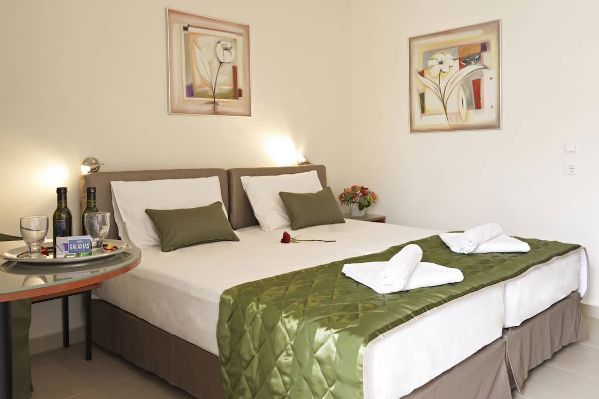 Quarto de hotel com cama, almofadas, mesa de cabeceira com um copo, garrafa e um vaso com flores.