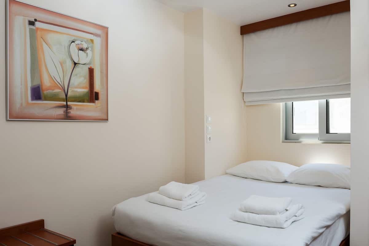 Quarto de hotel com cama, janela com persiana branca e um quadro na parede.