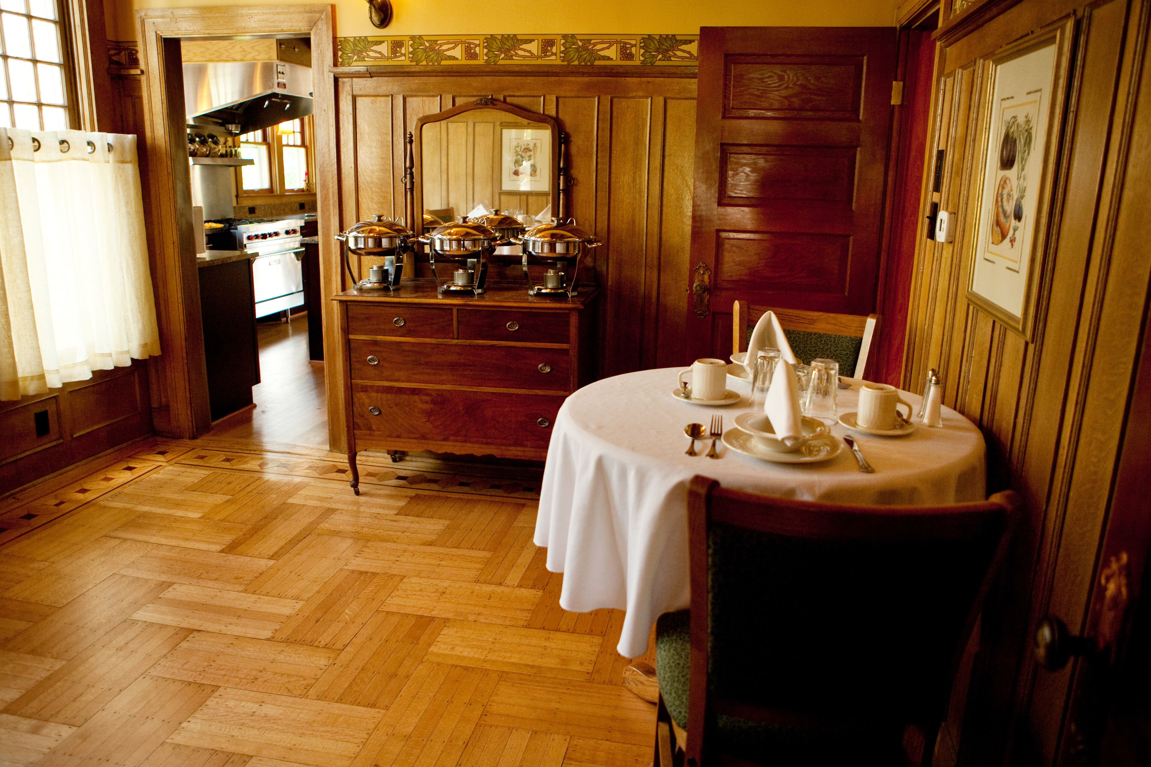 Uma área de refeições com uma mesa de madeira posta para quatro, um armário de madeira com panelas e uma cozinha ao fundo.