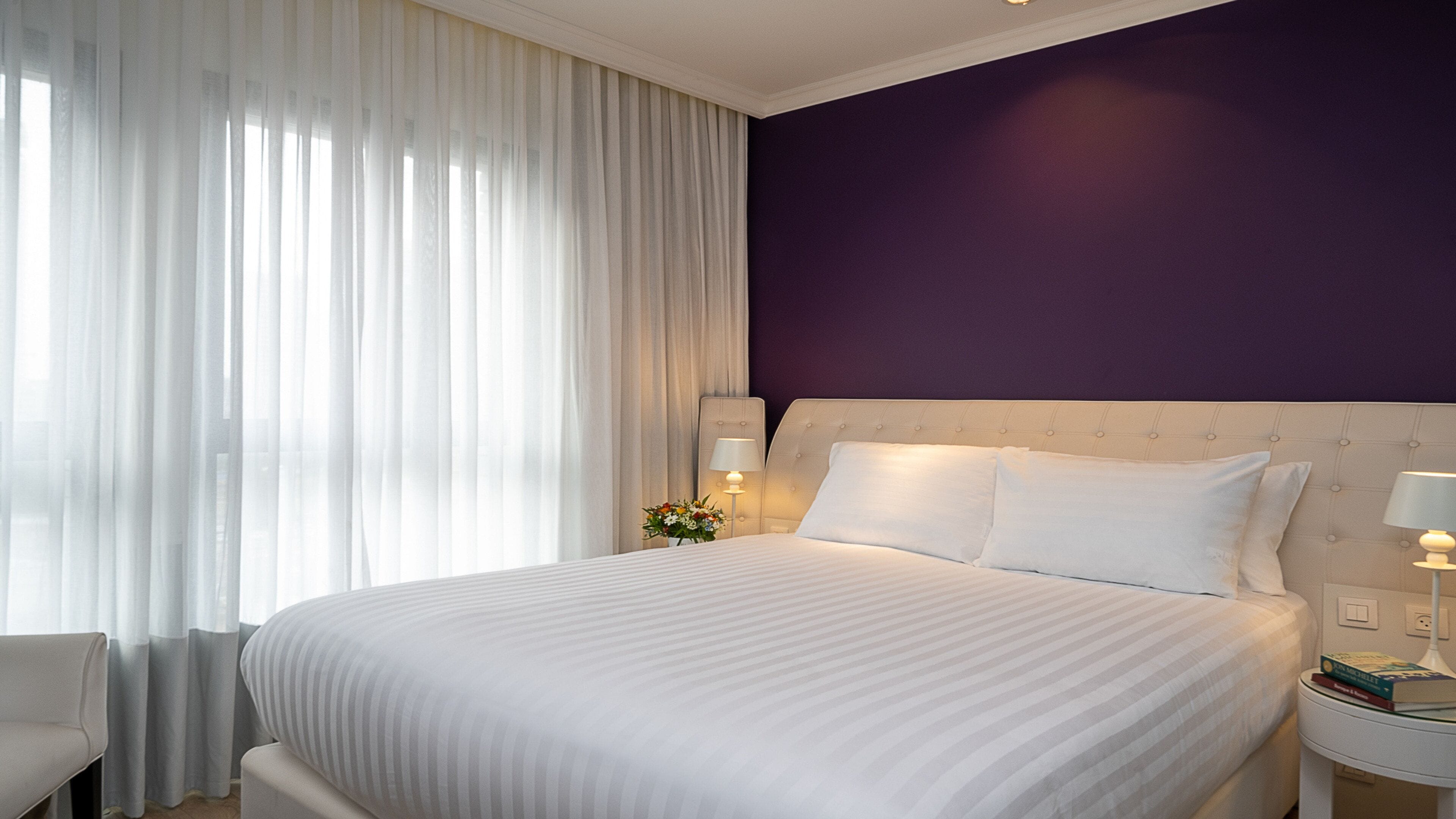 Quarto de hotel com uma cama grande, mesa de cabeceira com abajur e uma cadeira.