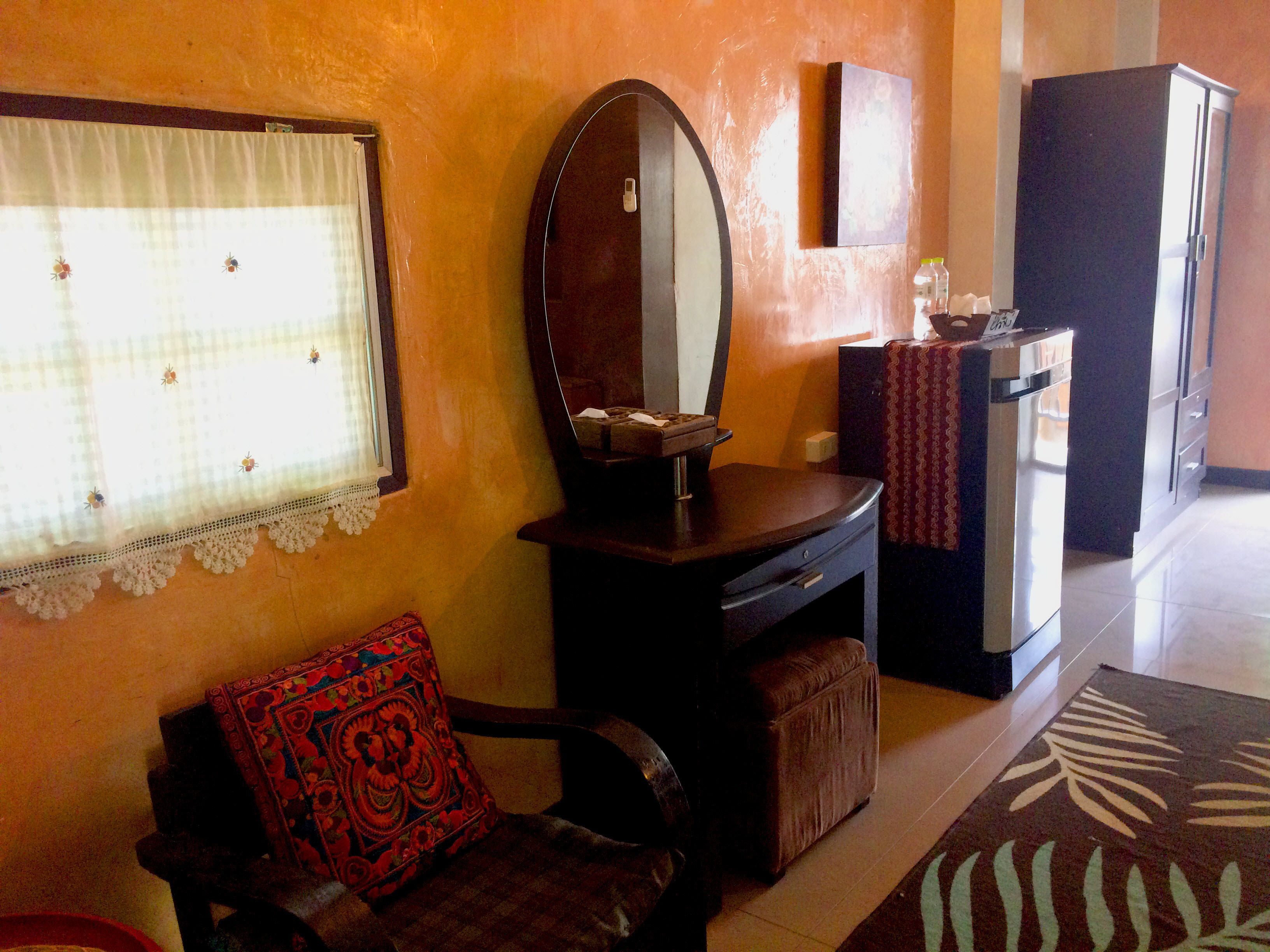 Quarto com cadeira de madeira, mesa pequena, espelho, geladeira e tapete estampado.