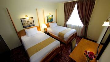 Quarto de hotel com duas camas de solteiro, piso de madeira, uma mesa de madeira com um vaso de flores e uma janela com cortinas.