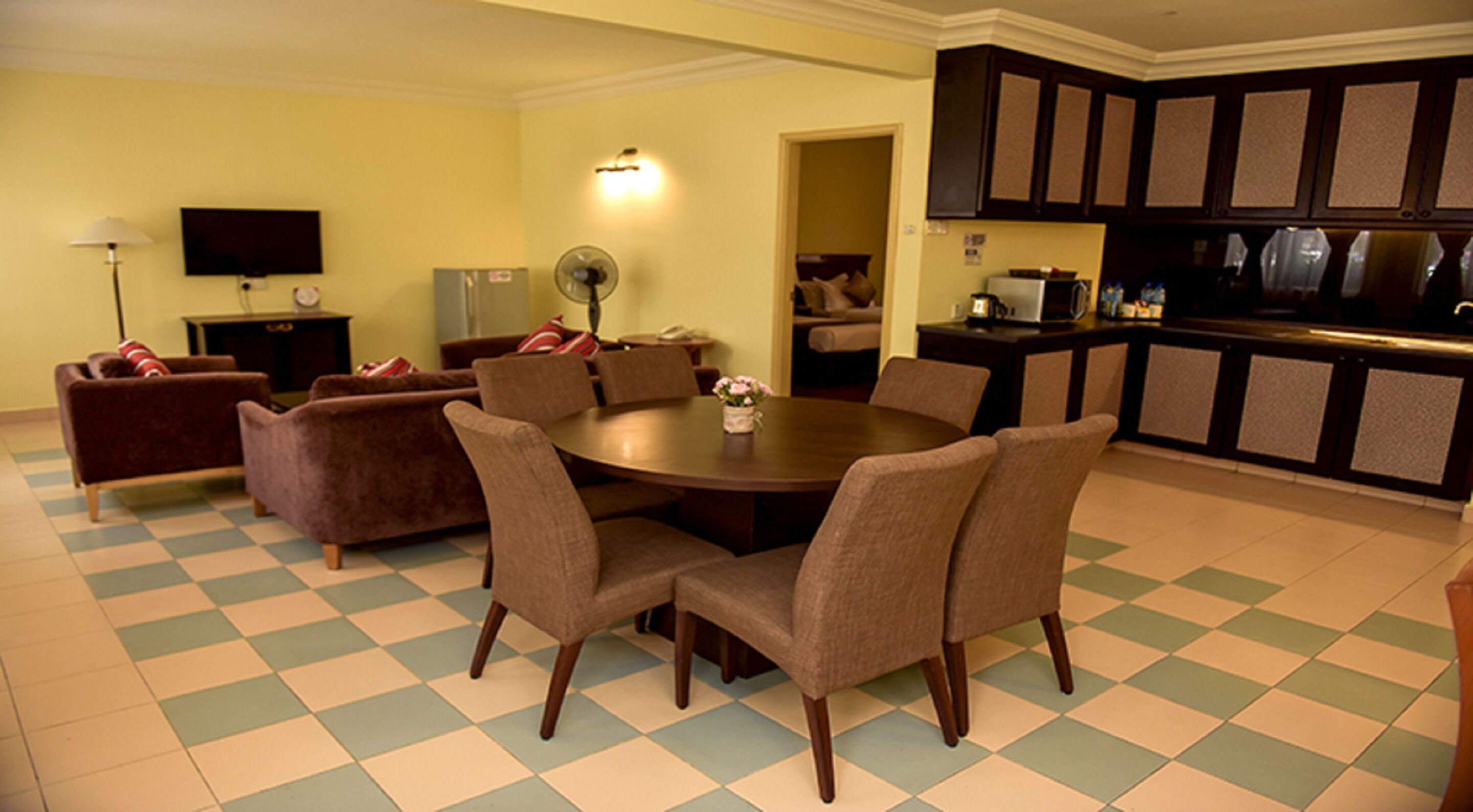 Uma sala de jantar com uma mesa redonda e seis cadeiras, uma televisão, um ventilador e uma cozinha com armários e eletrodomésticos.