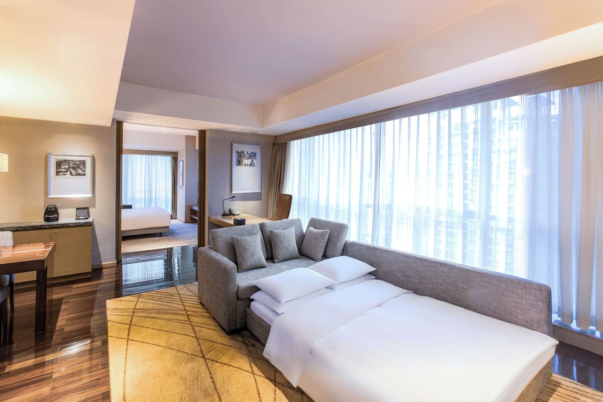 Quarto de hotel com uma cama grande, um sofá, uma mesa de escritório e vista para a cidade através de janelas amplas.