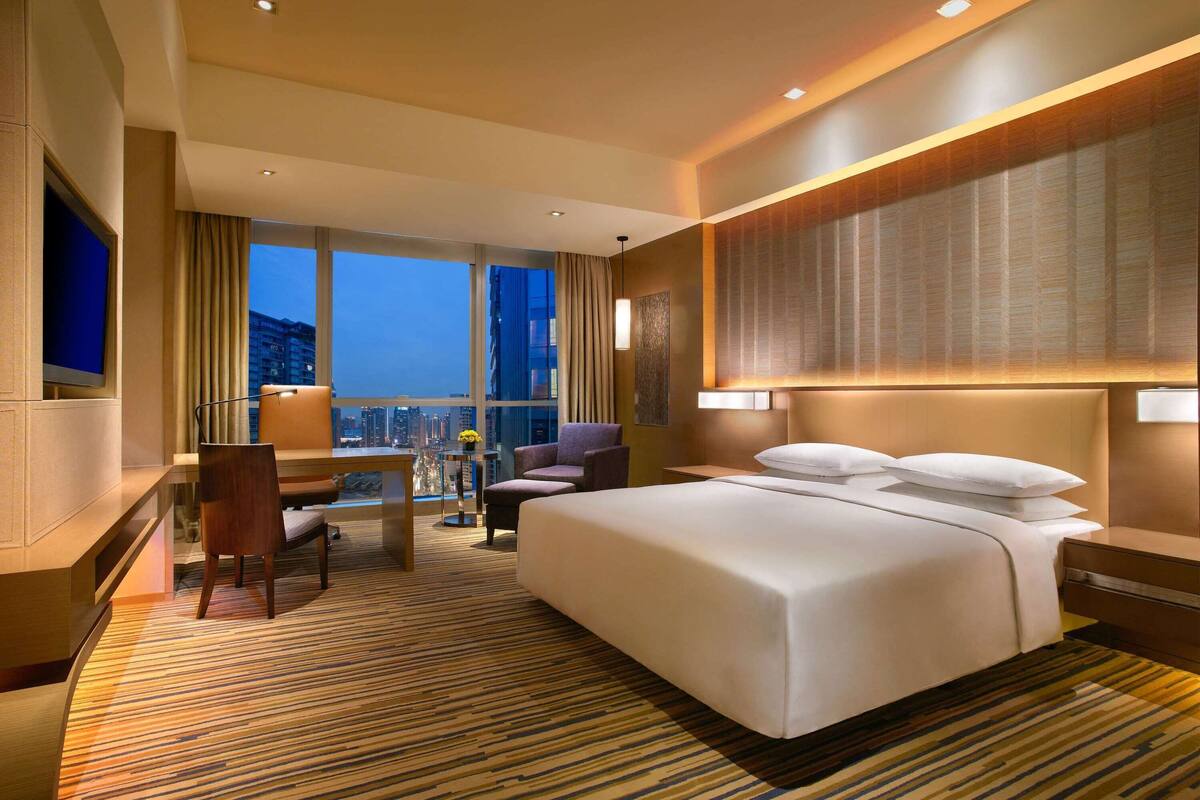 Quarto de hotel moderno com uma cama grande, uma escrivaninha, uma cadeira e vista para a paisagem urbana.