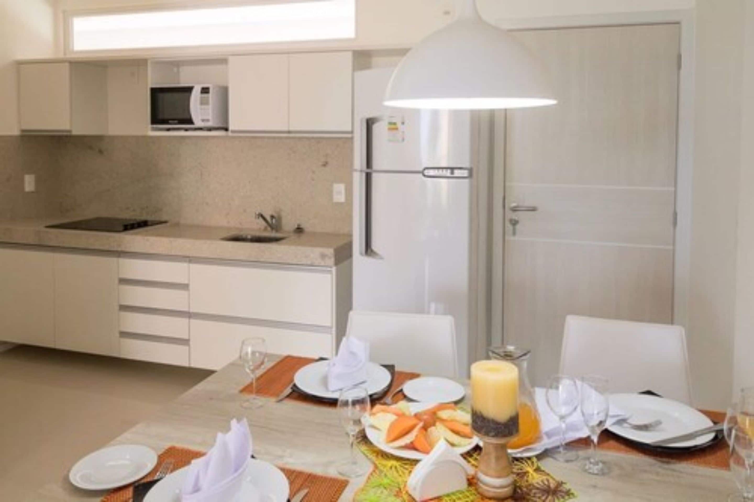 Cozinha moderna com armários brancos, micro-ondas e geladeira. Há uma mesa de jantar posta com pratos, copos e uma vela.
