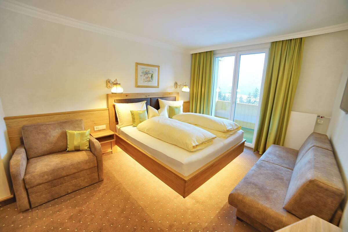 Quarto de hotel com uma cama grande, duas poltronas, uma cabeceira de madeira e uma janela com cortinas.
