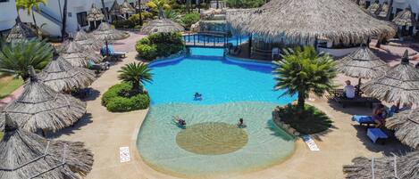 Área de piscina em resort, com piscina azul cristalina, guarda-sóis de palha e palmeiras.