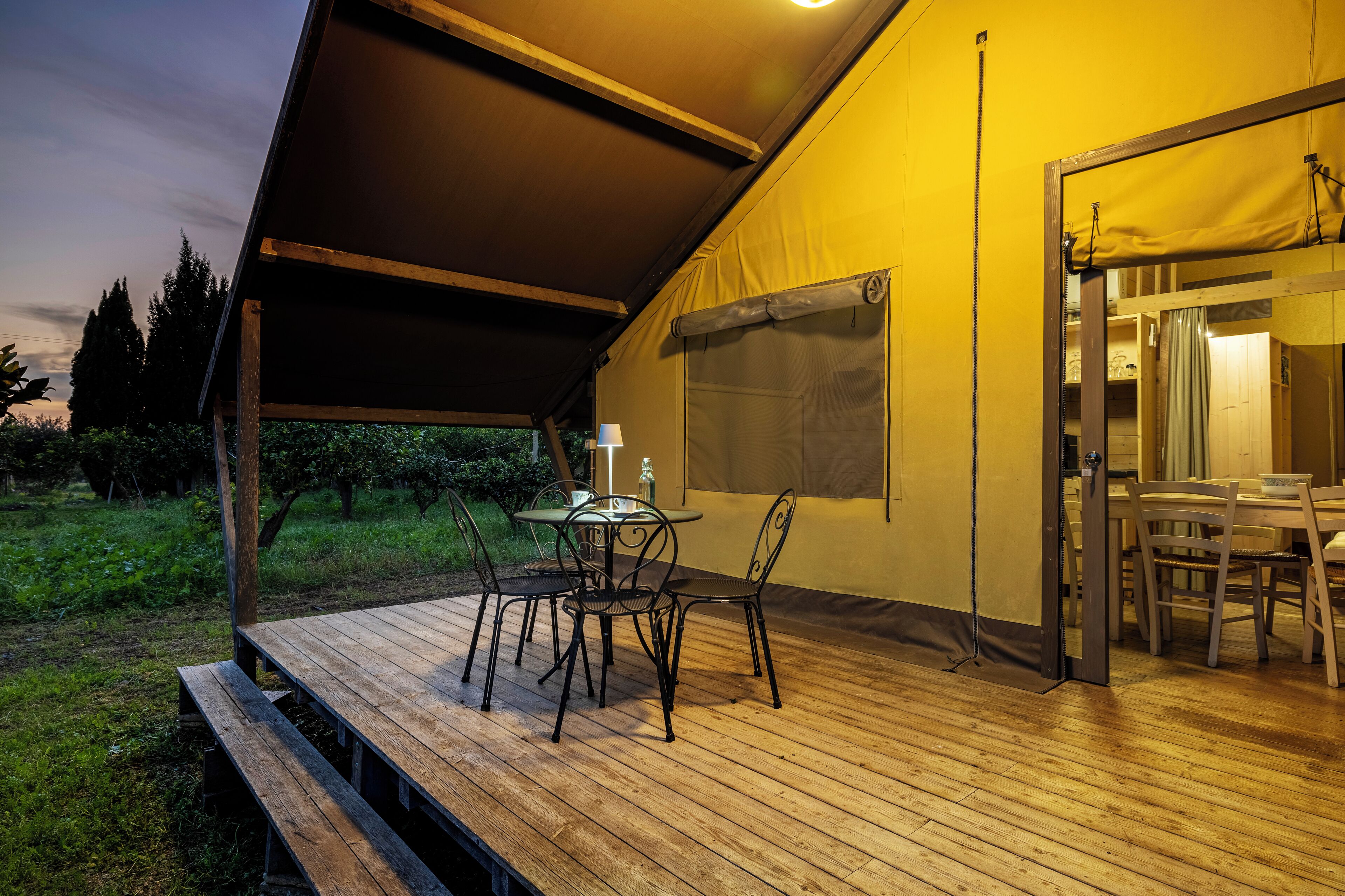 Uma tenda com deck de madeira, uma mesa com cadeiras e um abajur.