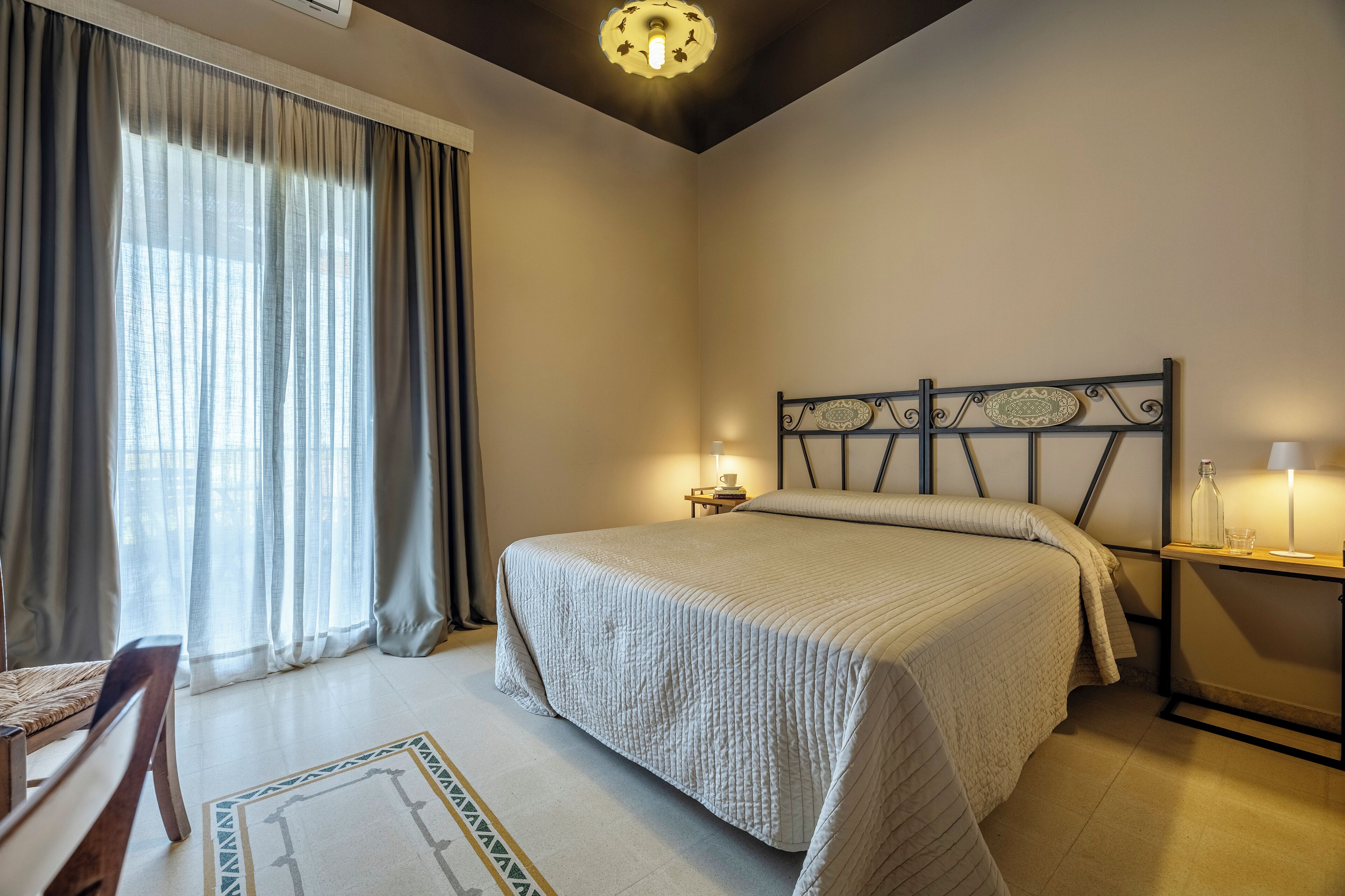 Quarto com cama, mesa de cabeceira, cadeira e janela com cortinas.
