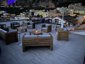 Terraço no telhado com mobiliário de madeira, painéis solares e vista para a paisagem urbana ao entardecer.