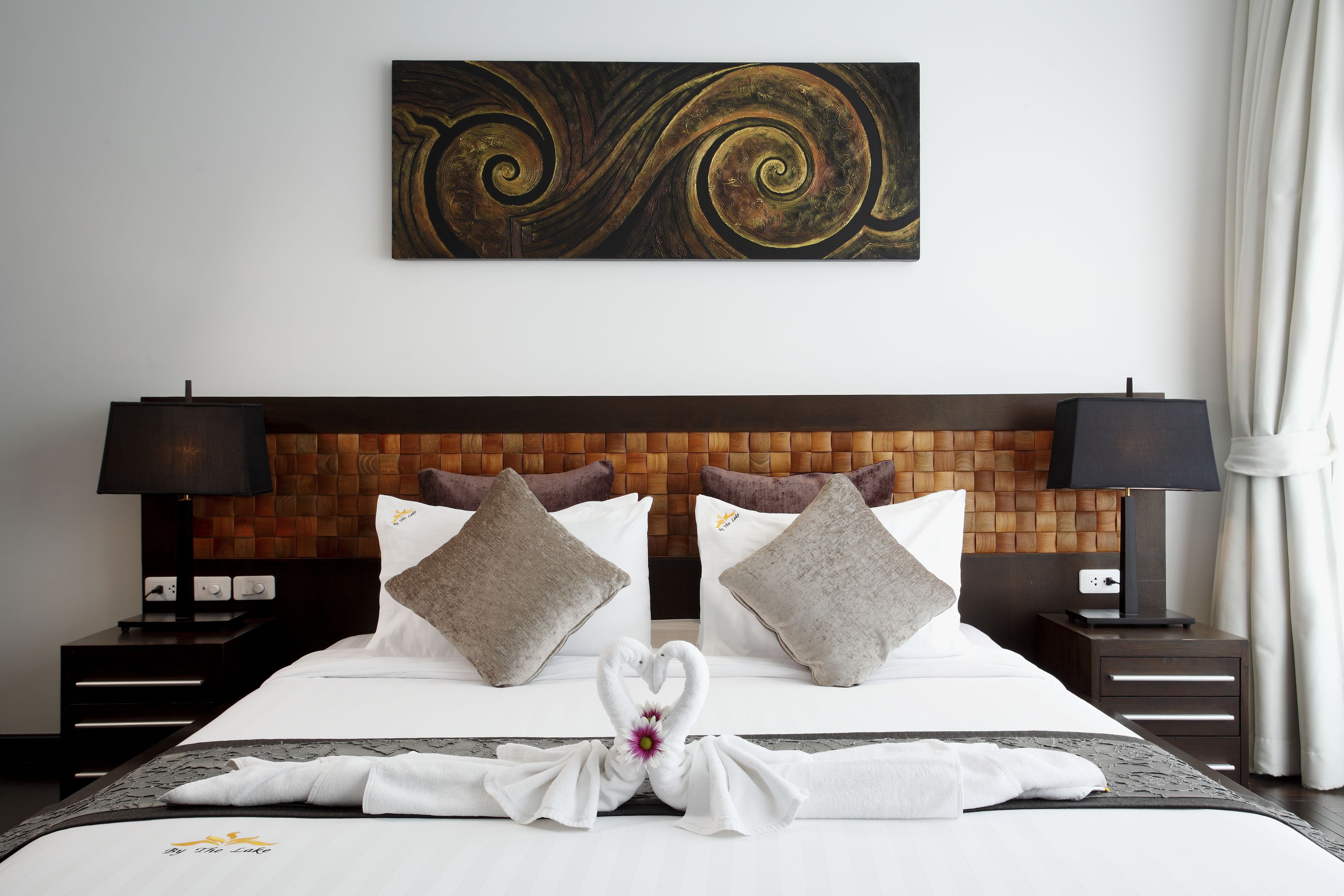 Quarto de hotel com uma cama bem arrumada, duas mesas de cabeceira com abajures, uma cabeceira com padrão trançado e um quadro abstrato grande na parede.