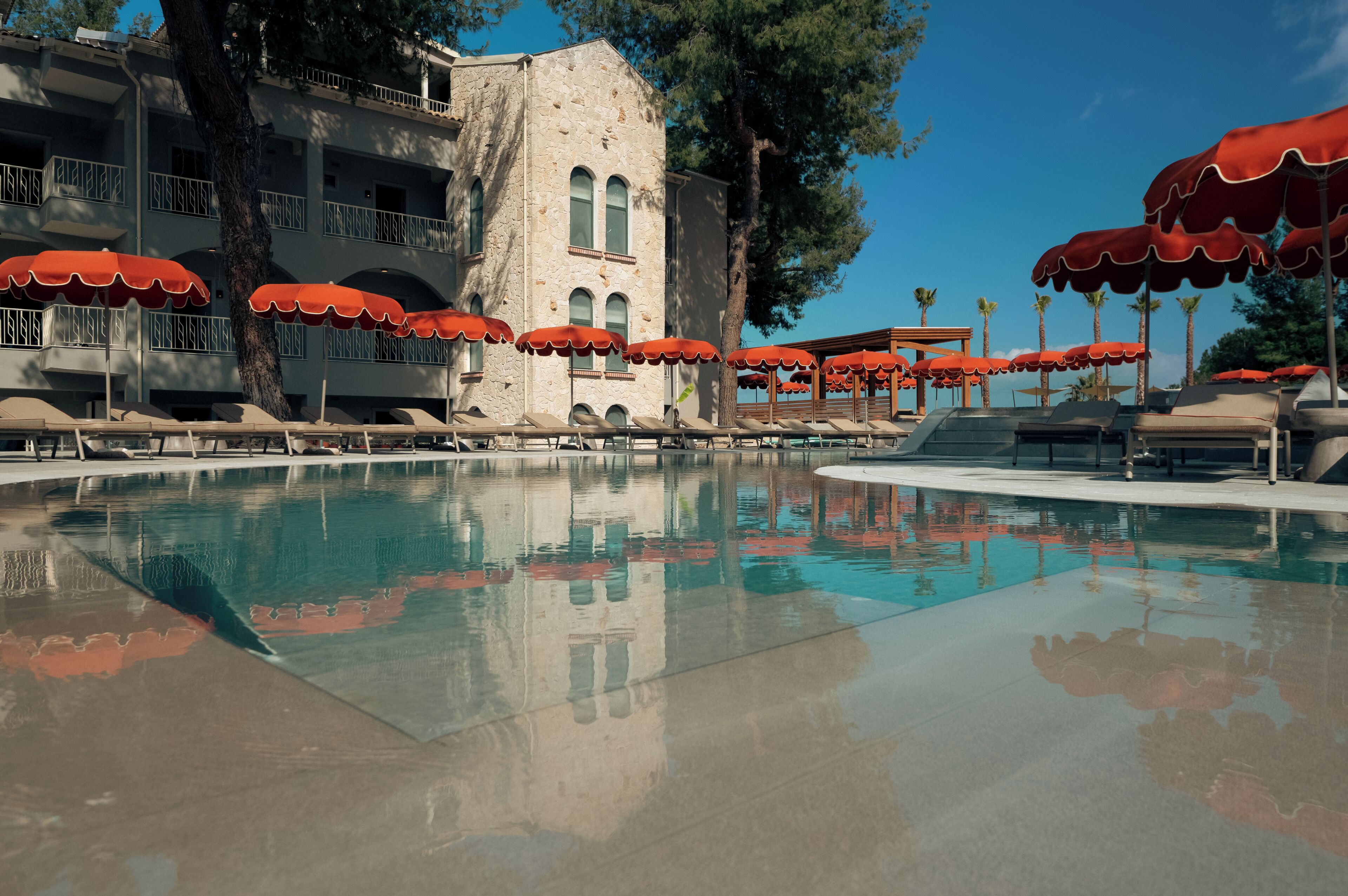 Piscina com guarda-sóis vermelhos, um edifício de pedra e céu claro.