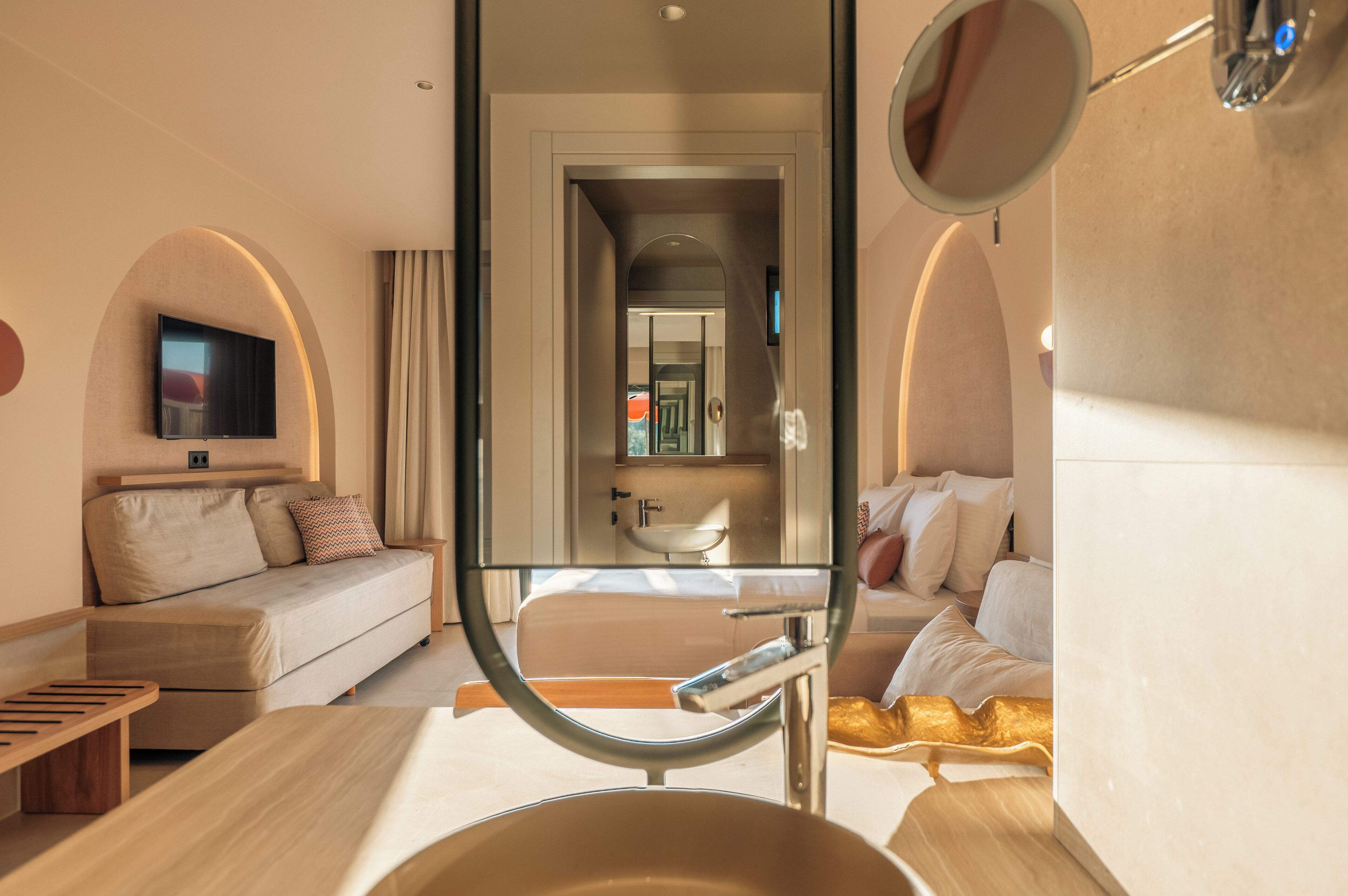 Quarto de hotel moderno com um espelho grande, um sofá, uma mesa de jantar e vista para o corredor.
