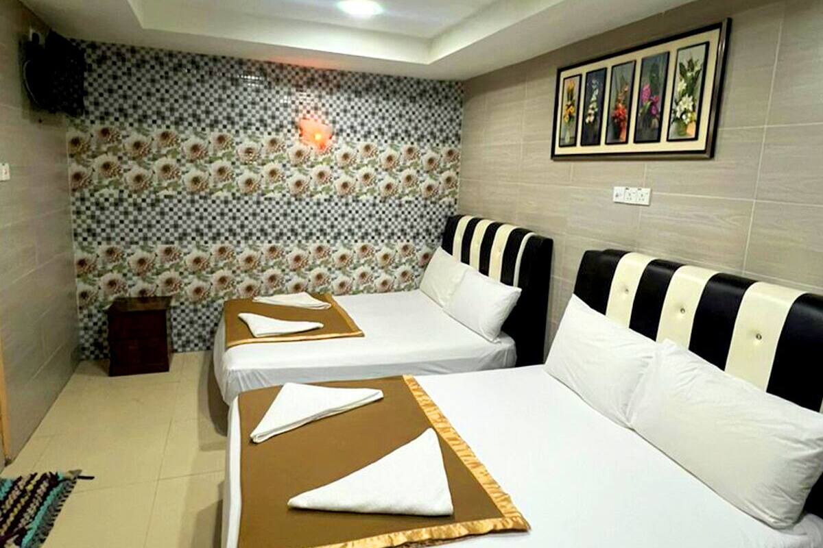 Quarto de hotel com duas camas, parede com padrão floral quadriculado, uma mesinha e quadros decorativos na parede.