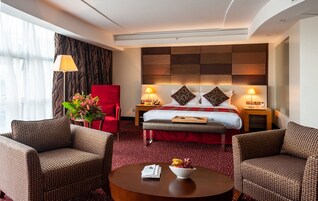 Quarto de hotel com uma cama grande, duas mesas de cabeceira com luminárias, uma poltrona vermelha, duas poltronas, uma mesa de centro redonda com uma tigela de frutas, e uma janela com cortinas leves.