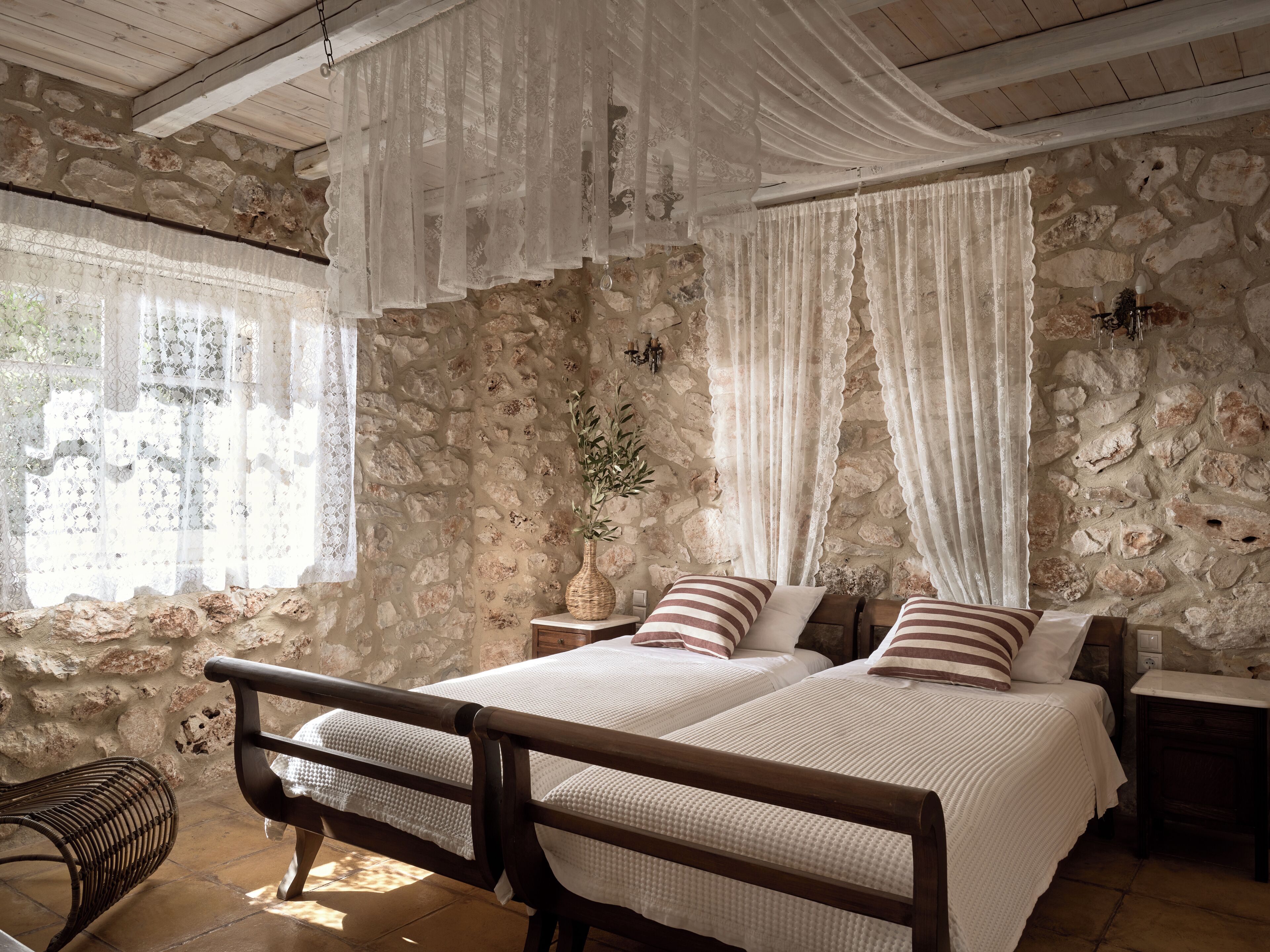 Quarto com duas camas, paredes de pedra e uma janela com cortinas de renda.