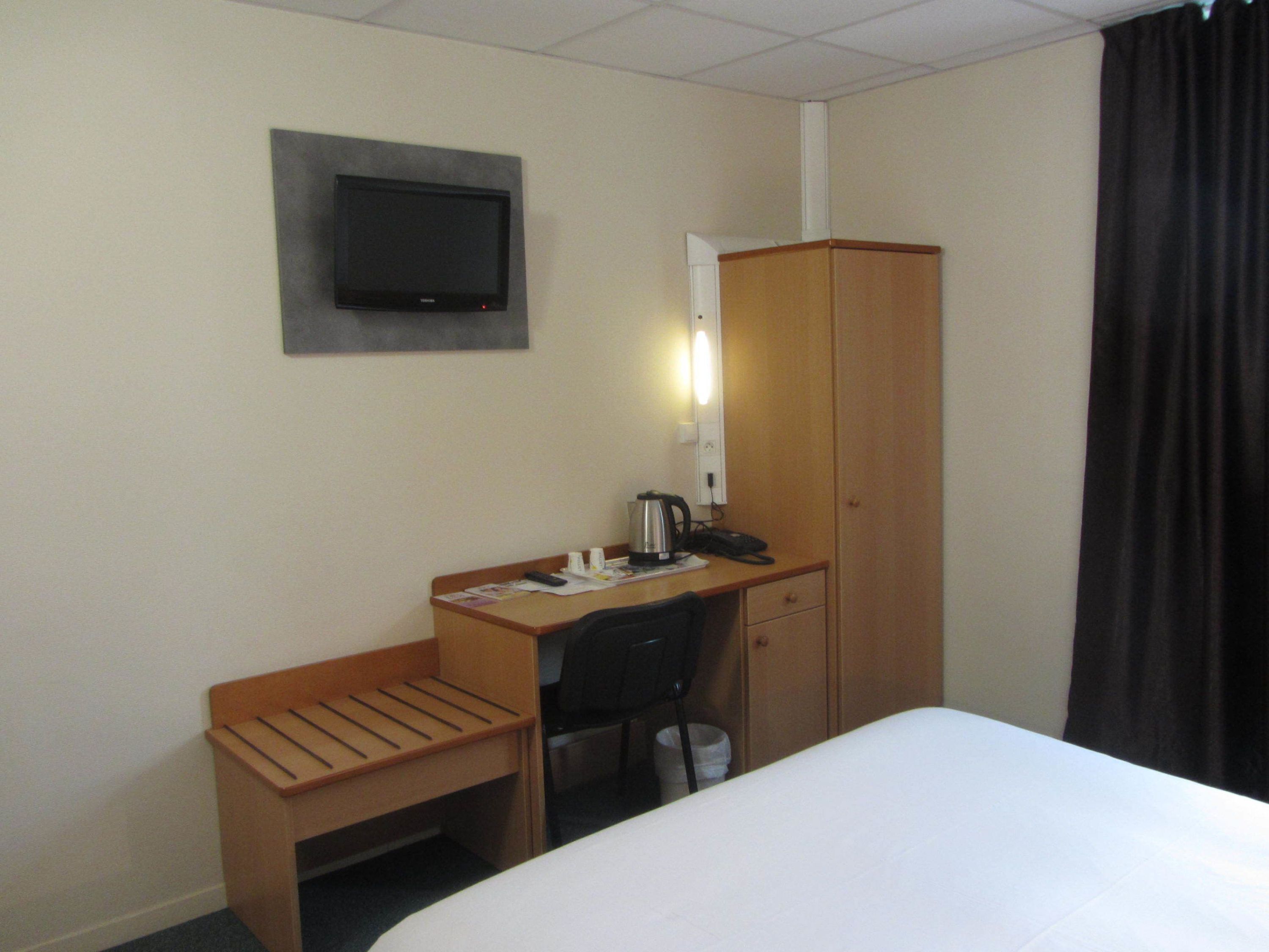 Quarto de hotel com uma cama, uma mesa com cadeira, uma televisão na parede e um guarda-roupa.