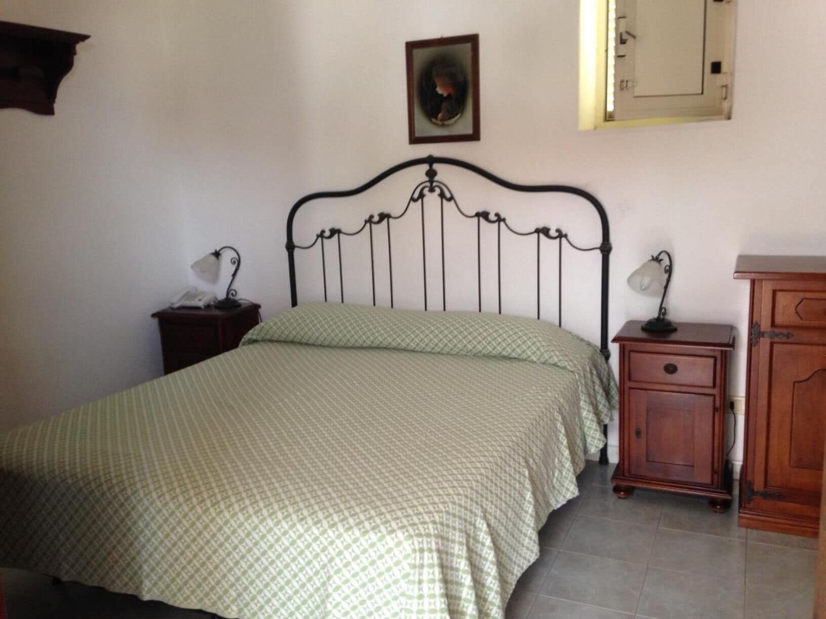 Quarto com estrutura de cama de metal, mesa de cabeceira com abajur, armário de madeira e quadro na parede.