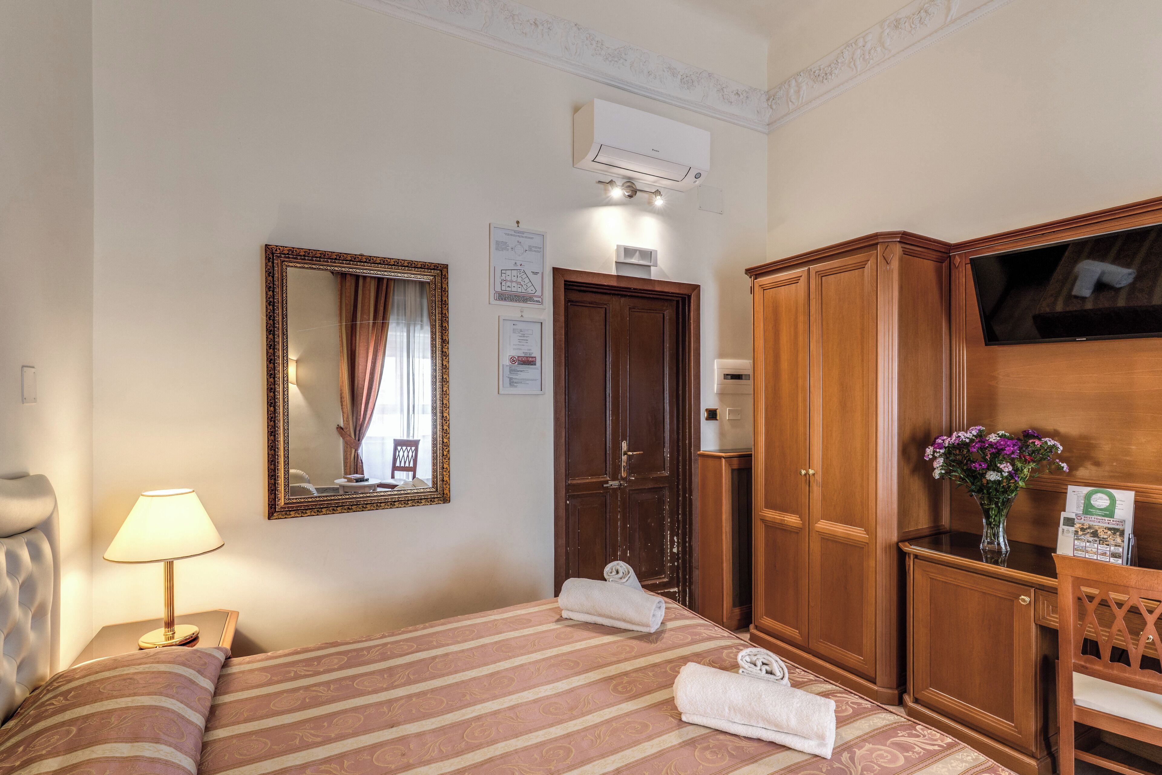 Quarto de hotel com cama, televisão, guarda-roupa de madeira, espelho e abajur.