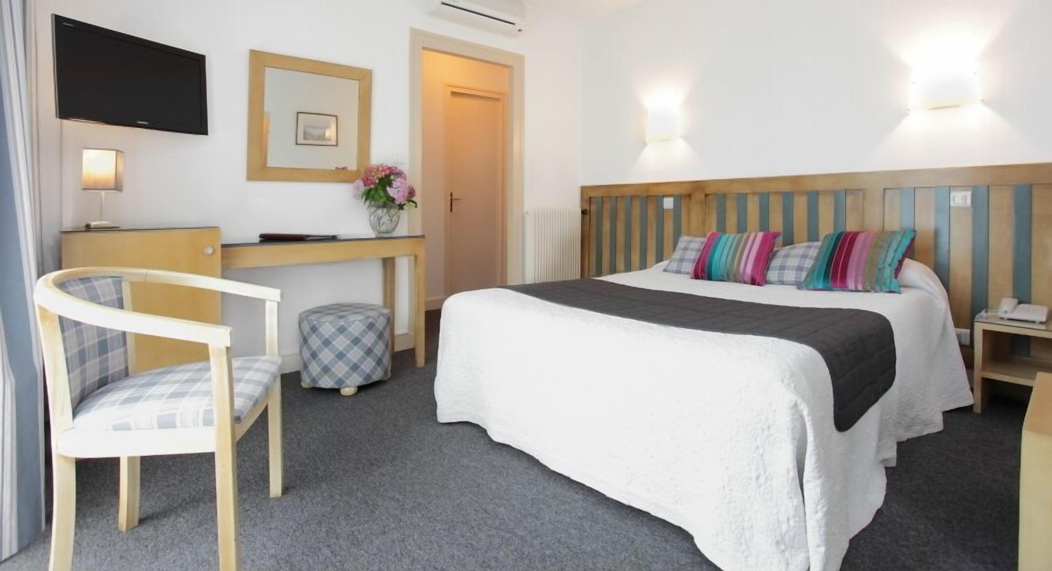Quarto de hotel com cama, escrivaninha, cadeira e televisão.