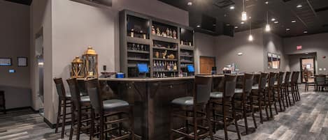 Uma área de bar com um balcão comprido, vários bancos altos e prateleiras com garrafas e copos.