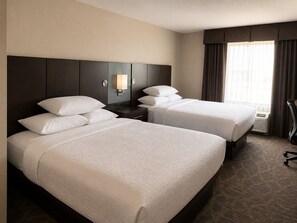 Quarto de hotel com duas camas, mesa de cabeceira, luminária, cadeira e janela com cortinas.