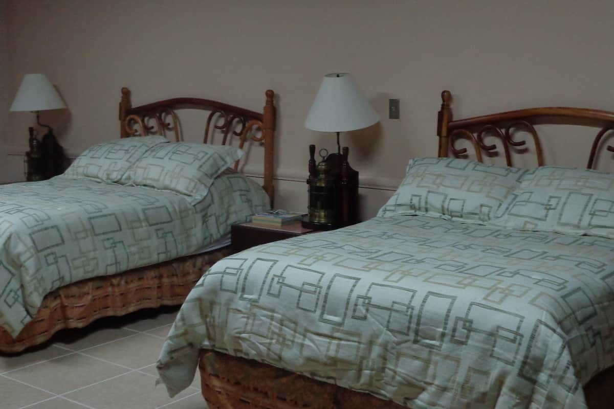 Duas camas de solteiro com cabeceiras de madeira, cada uma com uma mesa de cabeceira e uma luminária. As camas possuem roupa de cama estampada.
