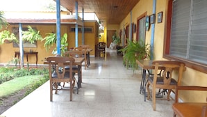 Uma área de estar coberta ao ar livre, com mesas e cadeiras de madeira, plantas em vasos e piso de azulejos.