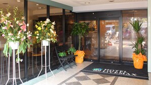 Entrada de hotel com piso xadrez, plantas em vasos e um banco.