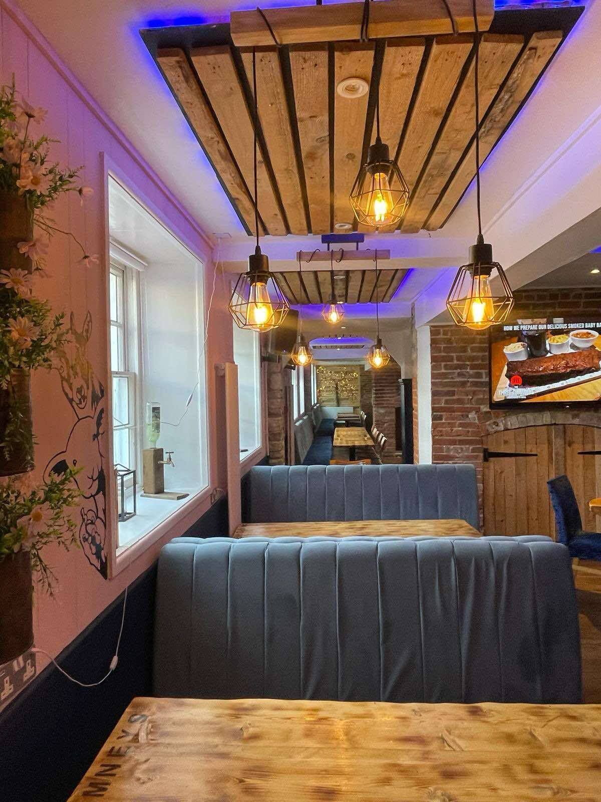 Interior de restaurante com vigas de madeira no teto, luminárias pendentes e área de estar com bancos acolchoados.