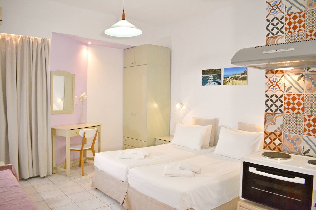 Quarto de hotel com duas camas, uma escrivaninha, uma cadeira, um guarda-roupa e uma cozinha compacta.