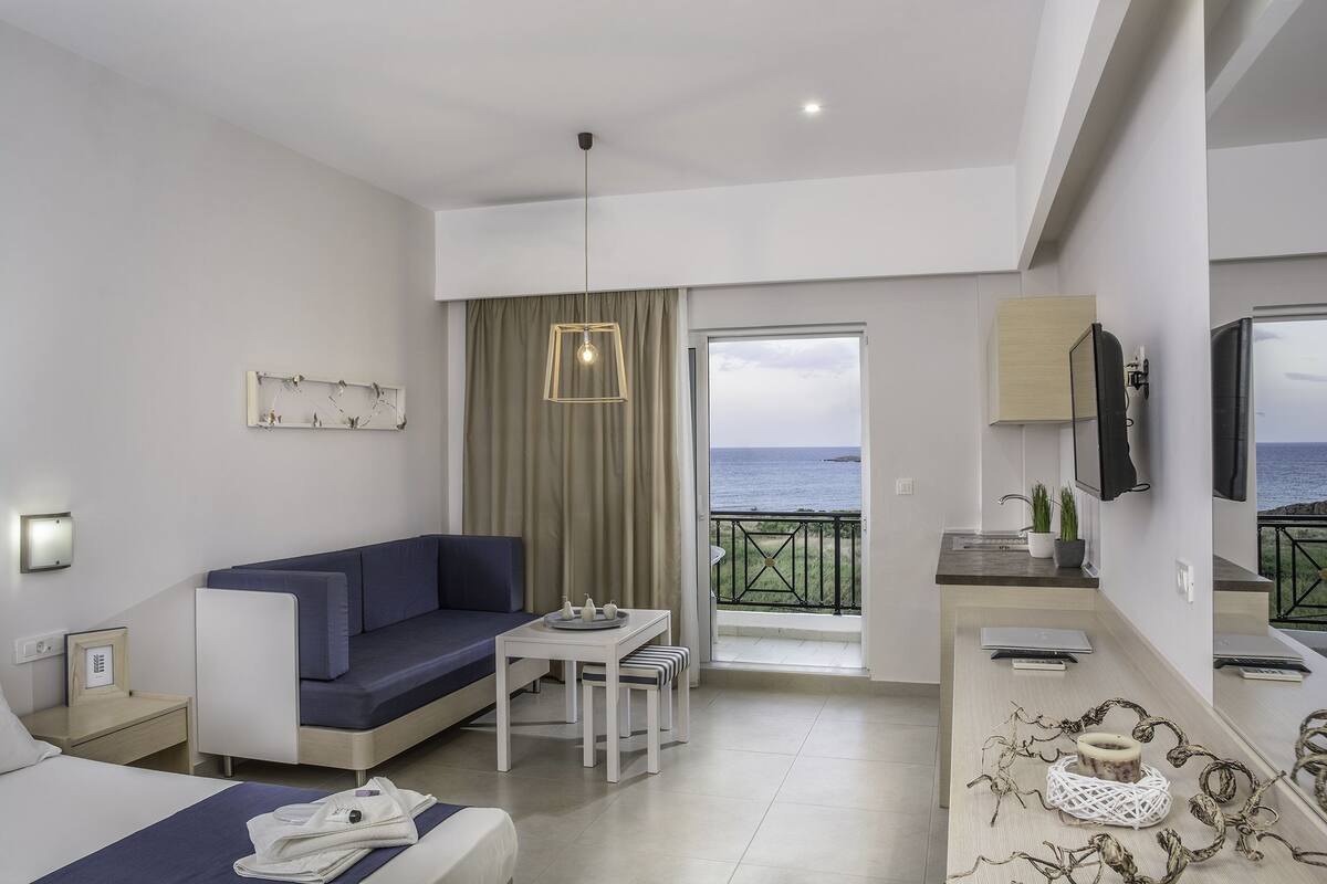 Quarto de hotel moderno com sofá, mesa pequena, escrivaninha com laptop, televisão e varanda com vista para o mar.