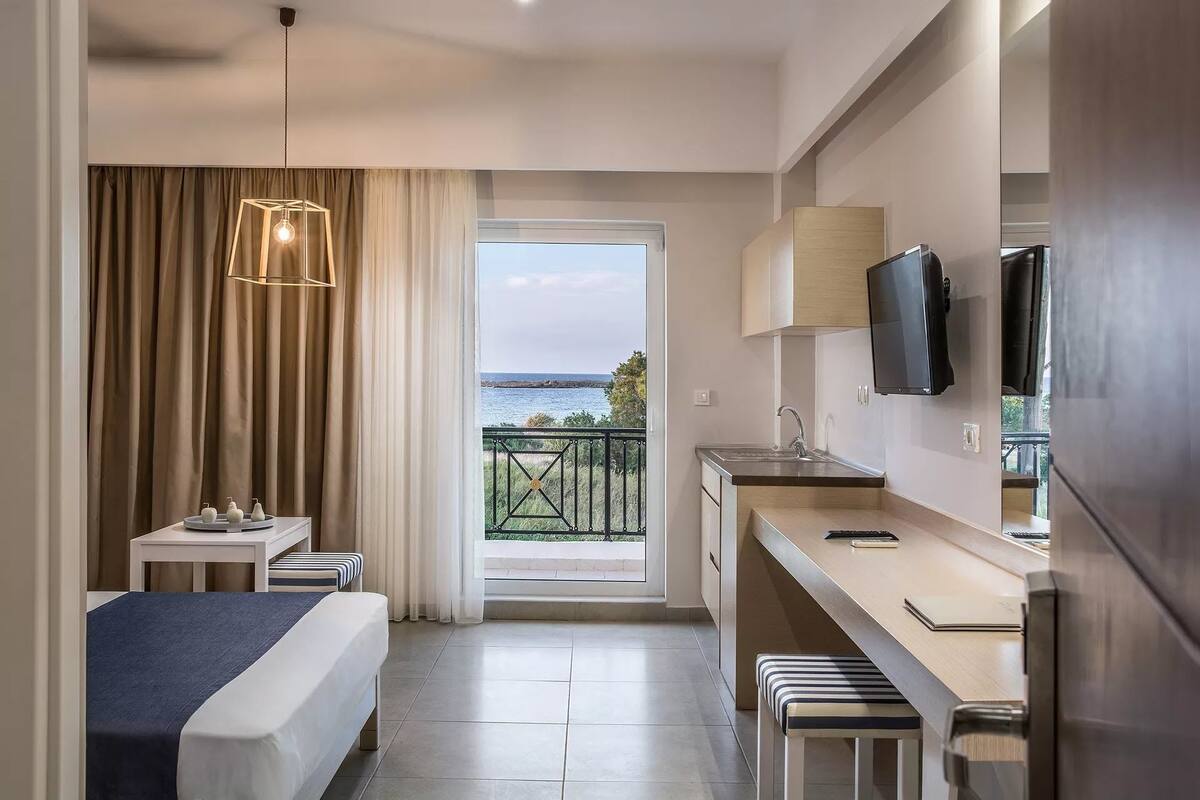 Quarto de hotel com cama, uma mesinha com produtos de higiene, uma escrivaninha com cadeira, televisão e varanda com vista para o mar.