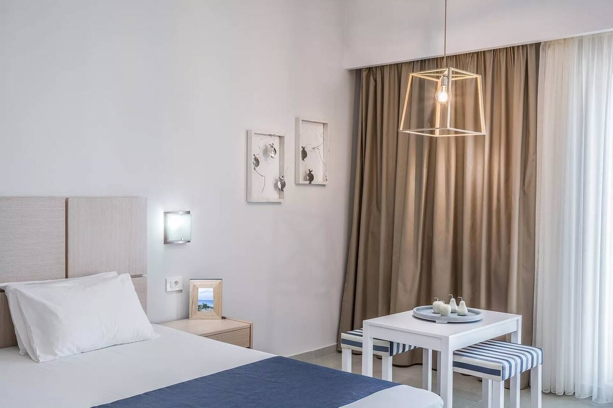 Quarto de hotel moderno com cama, mesa de cabeceira, criado-mudo e uma pequena mesa com bandeja.