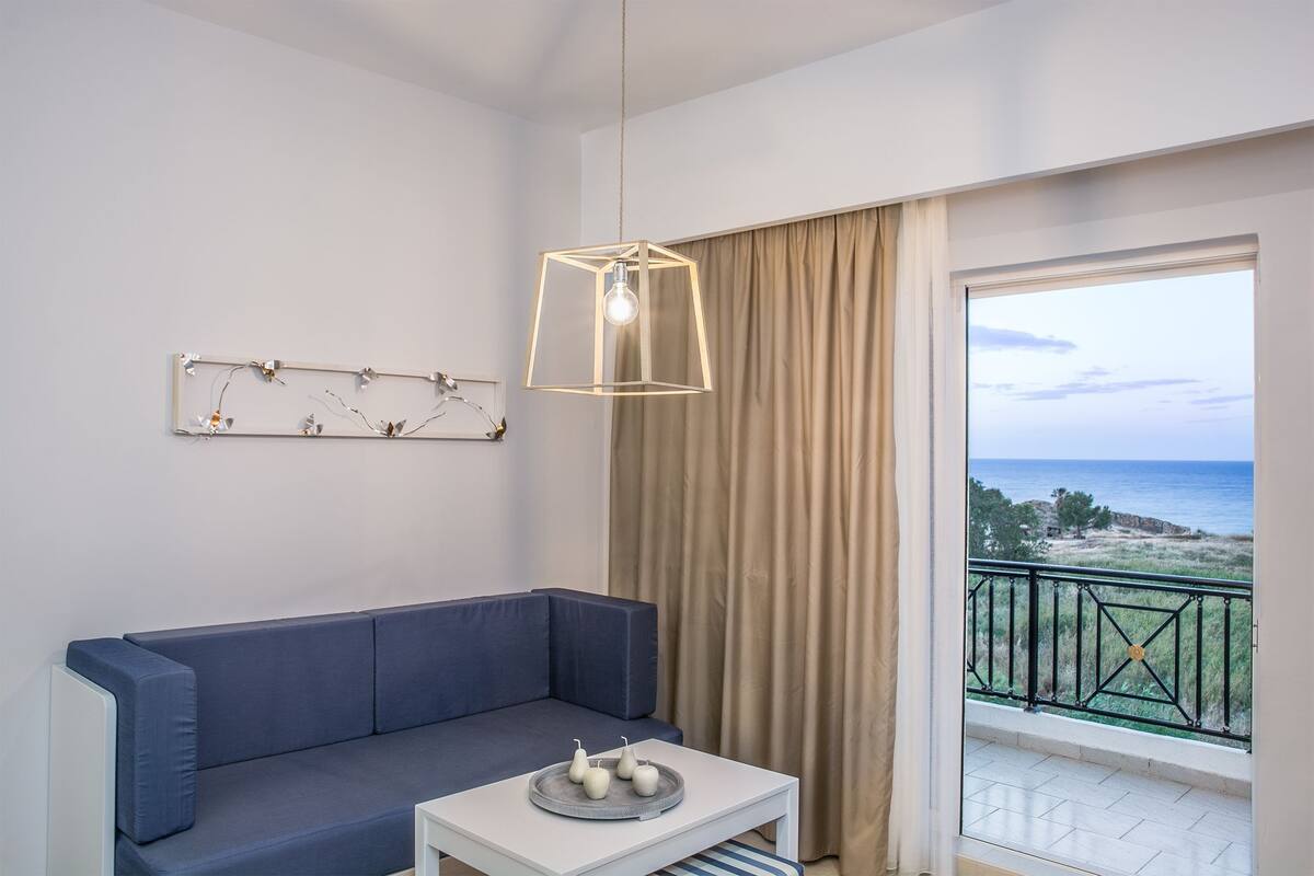 Uma sala de estar moderna com um sofá azul, uma mesa de centro branca com objetos decorativos, uma luminária pendente geométrica e vista para o mar através de uma varanda com guarda-corrimão preto.
