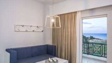 Uma sala de estar moderna com um sofá azul, uma mesa de centro branca com objetos decorativos, uma luminária pendente geométrica e vista para o mar através de uma varanda com guarda-corrimão preto.