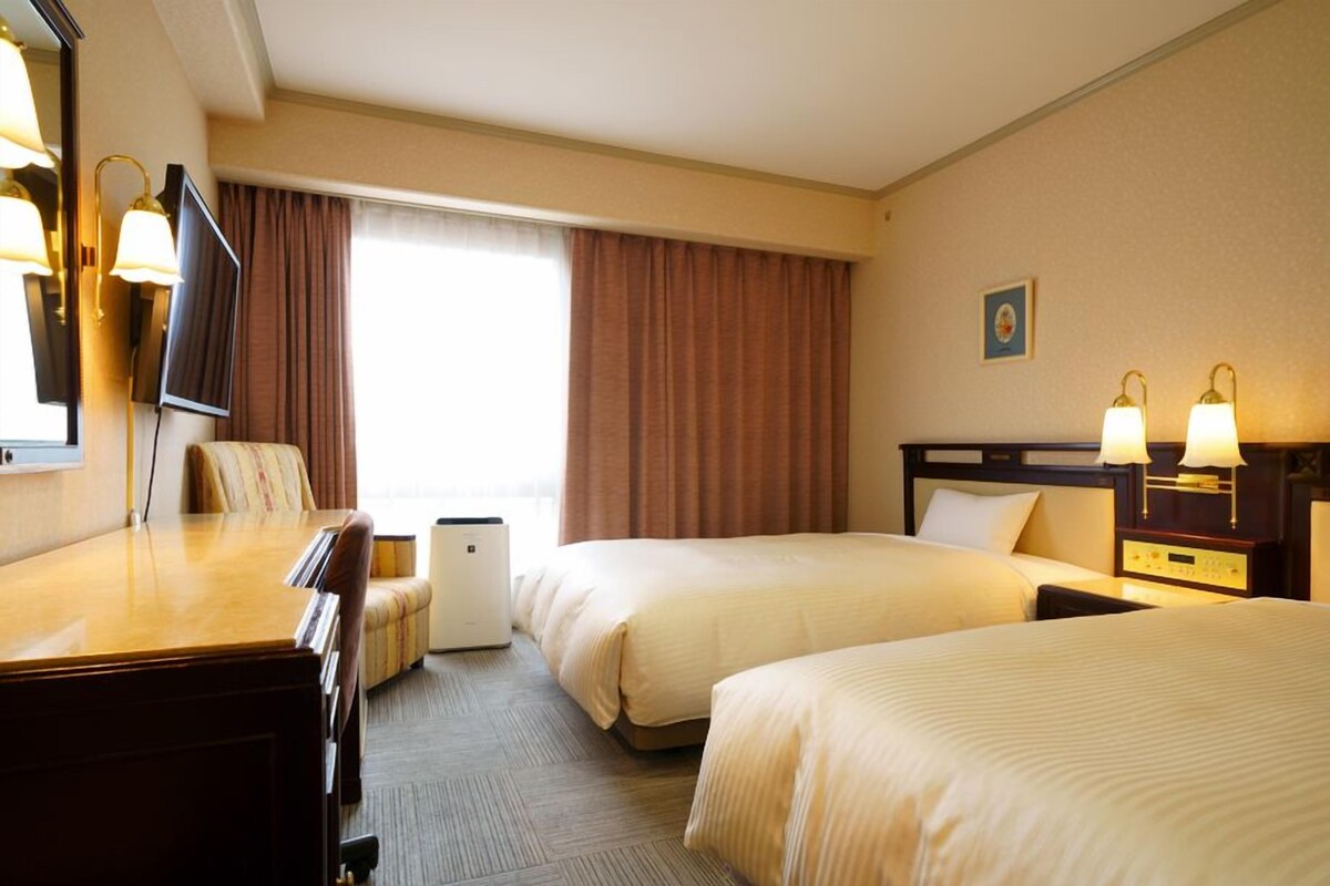 Quarto de hotel com duas camas, uma escrivaninha, televisão e janela com cortinas.