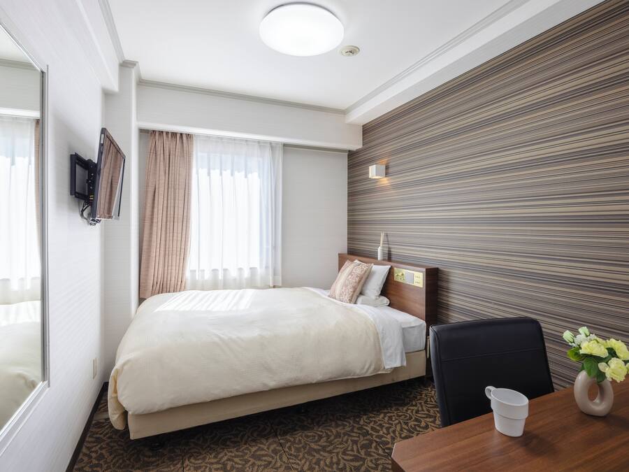Quarto de hotel com uma cama, uma escrivaninha com cadeira, televisão fixada na parede e uma janela com cortinas.