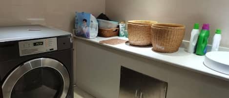 Área de lavanderia com máquina de lavar, cestos e produtos de limpeza.