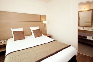 Quarto de hotel com uma cama grande, uma mesa de cabeceira, abajur, espelho e banheiro com porta-toalhas.