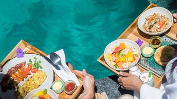 Duas pessoas jantando em mesa de madeira perto de uma piscina com água cristalina, pratos de comida e bebidas.