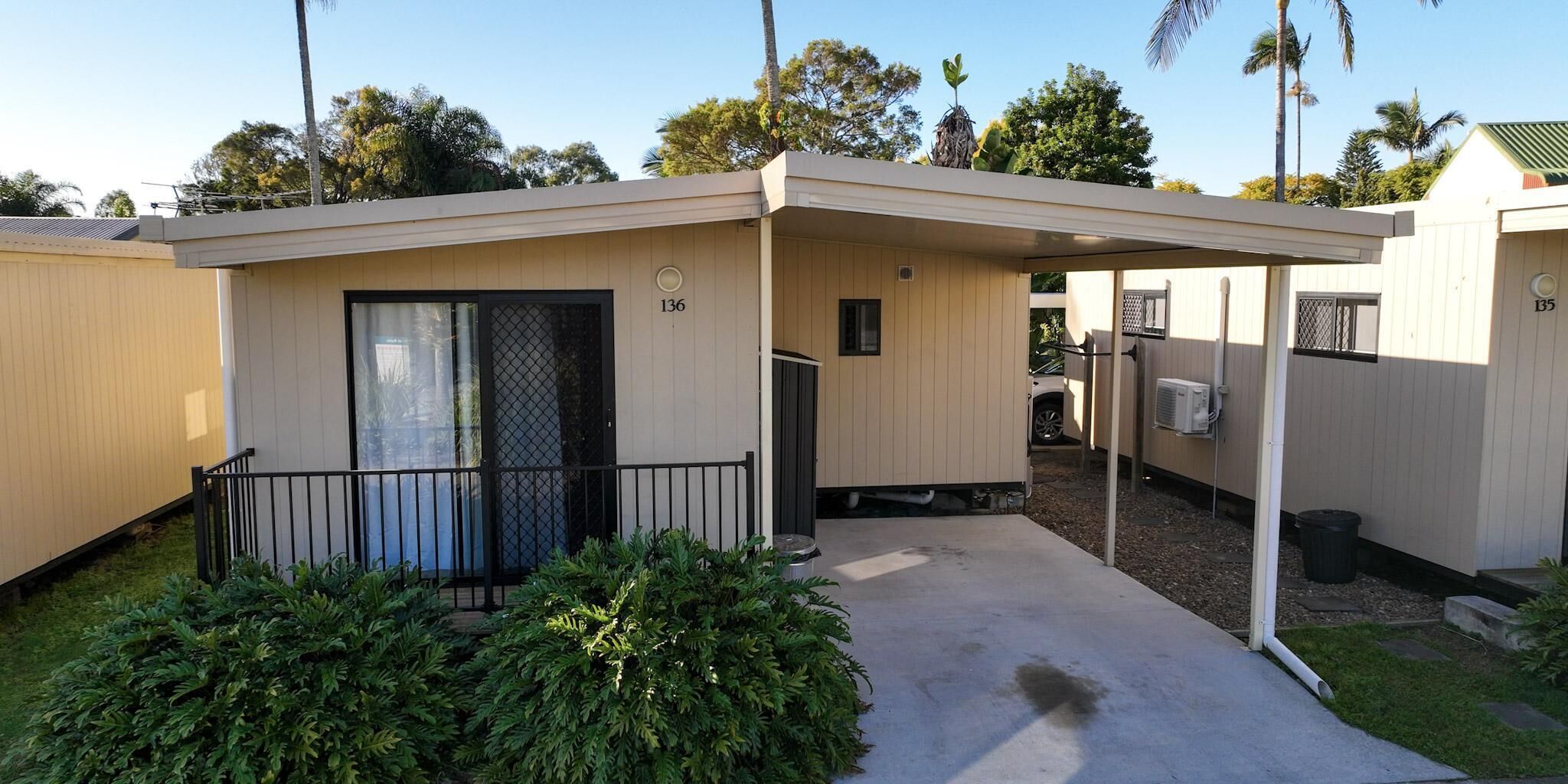Uma fileira de casas móveis térreas, com portas numeradas, uma área externa coberta e um pequeno jardim.