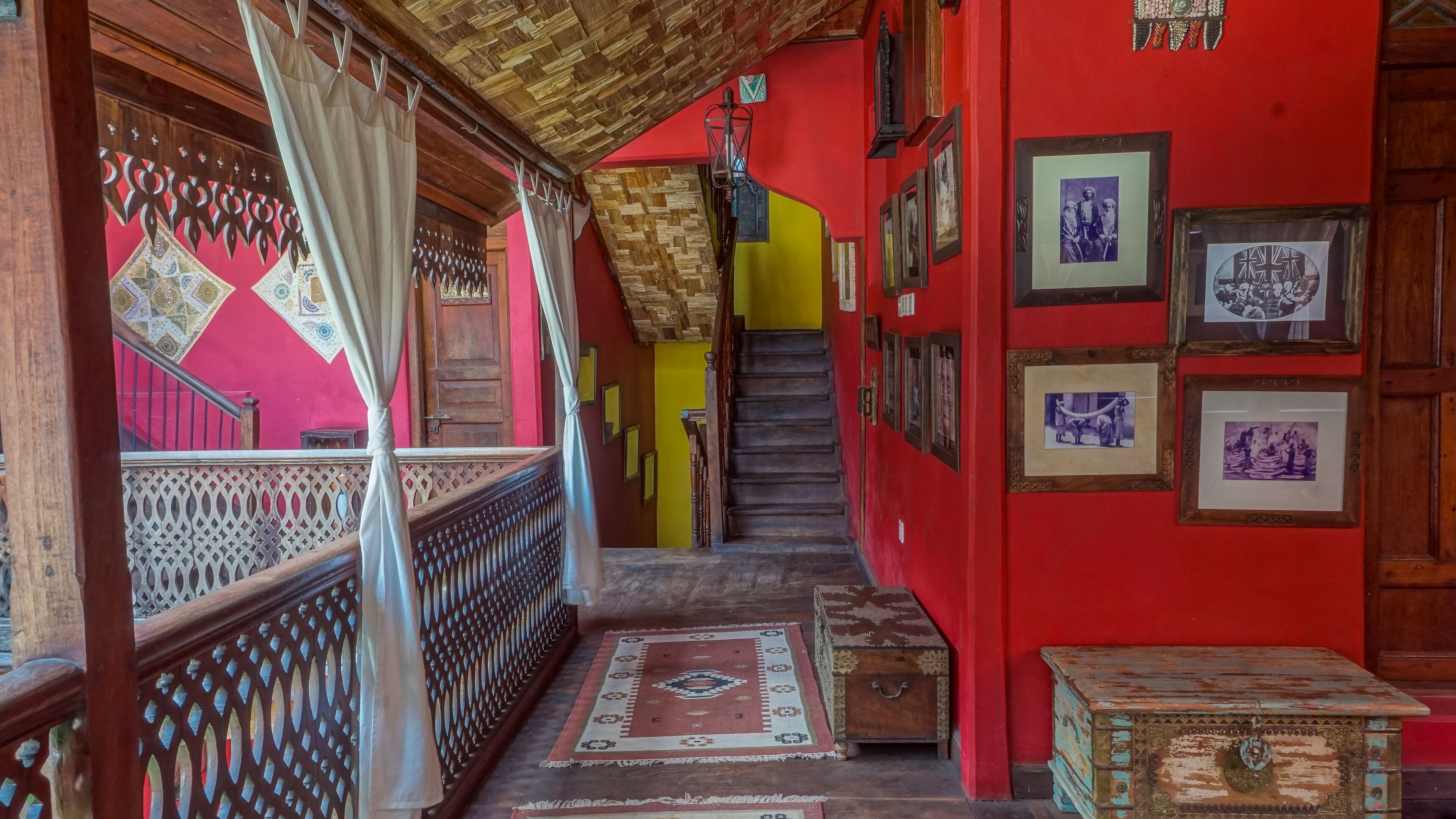 Uma escadaria com corrimão de madeira, uma parede vermelha com quadros e um tapete estampado.