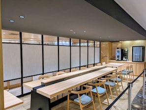Uma área de refeições moderna com longas mesas e cadeiras de madeira, uma cozinha com frigorífico e máquina de vendas, e um divisório com vista para o interior.
