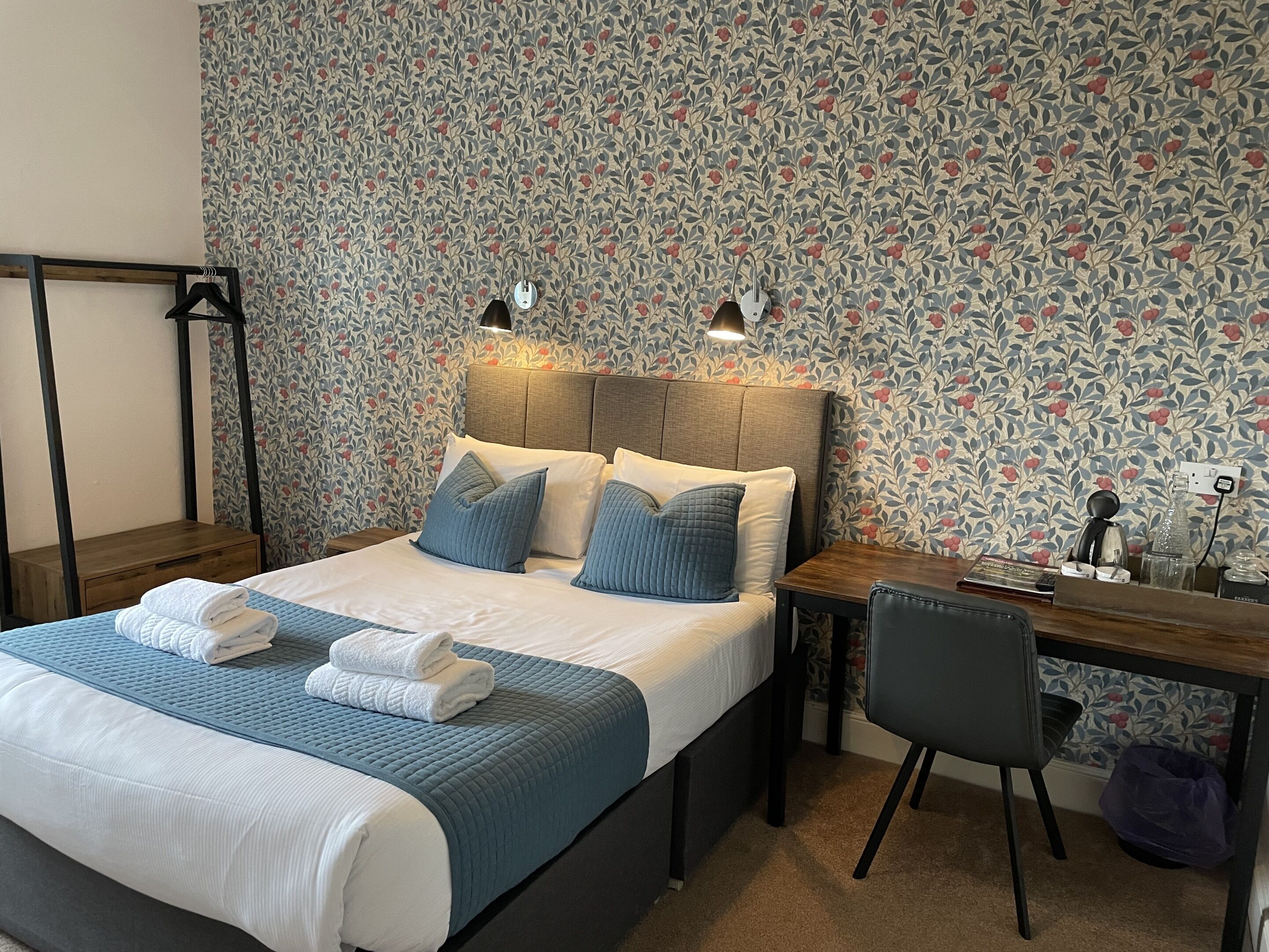 Uma cama bem arrumada com roupa de cama azul e branca, uma escrivaninha com cadeira e uma parede com padrão floral.